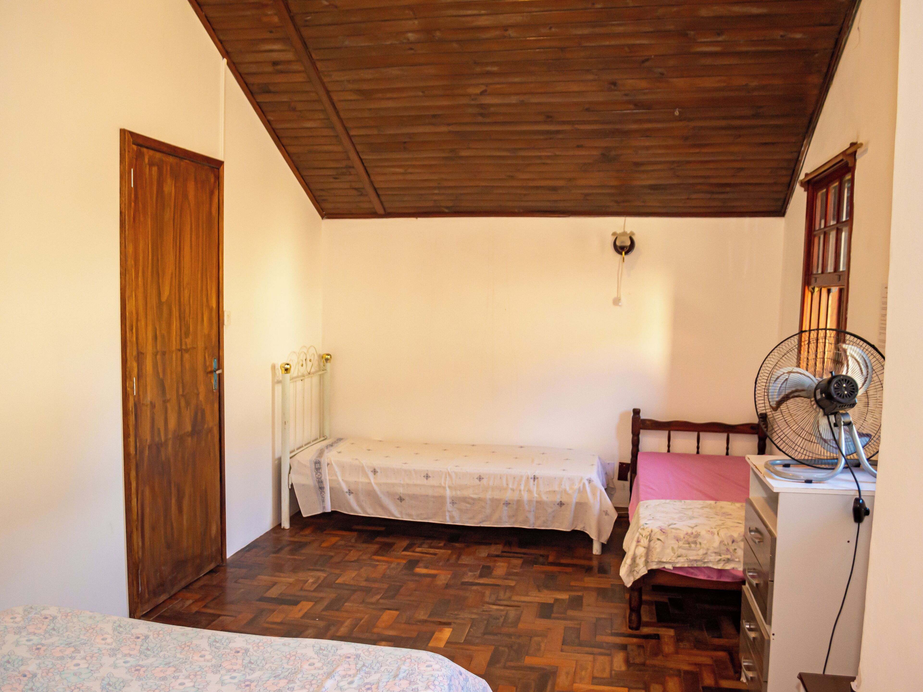 Quarto com teto de madeira, duas camas, radiador, porta de madeira e ventoinha.