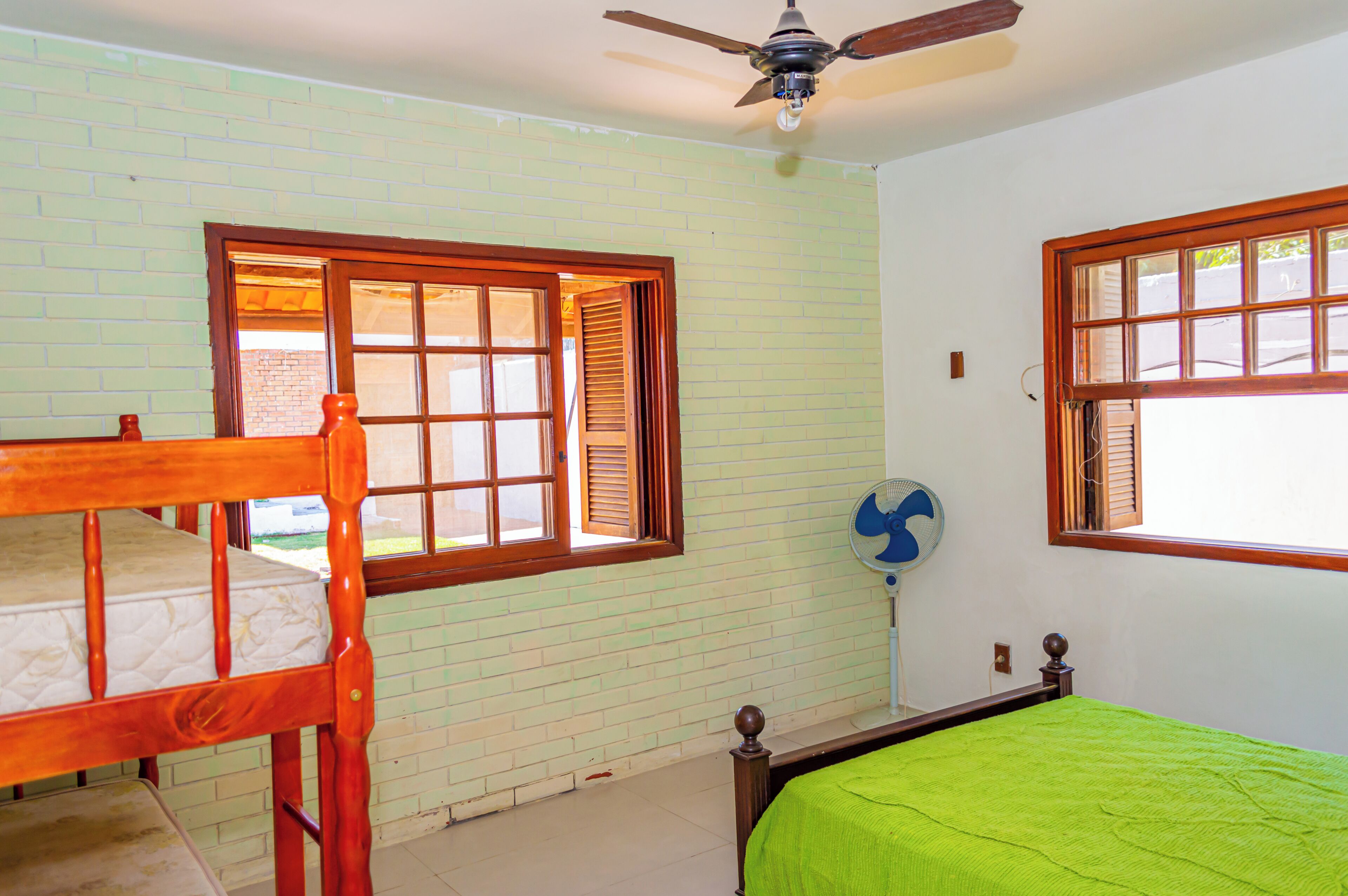 Quarto com cama, cadeira de madeira, ventilador e janela com persianas.