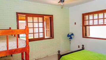 Quarto com cama, cadeira de madeira, ventilador e janela com persianas.