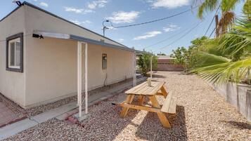 Um edifício residencial com um alpendre, uma mesa de madeira e um pequeno jardim com palmeiras.