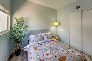 Um quarto com cama, mesa de cabeceira, abajur, planta em vaso e guarda-roupa.