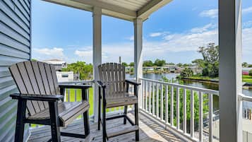 Um deck de madeira com duas cadeiras Adirondack e uma mesa, com vista para um corpo de água e uma área residencial.