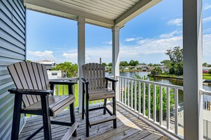 Um deck de madeira com duas cadeiras Adirondack e uma mesa, com vista para um corpo de água e uma área residencial.