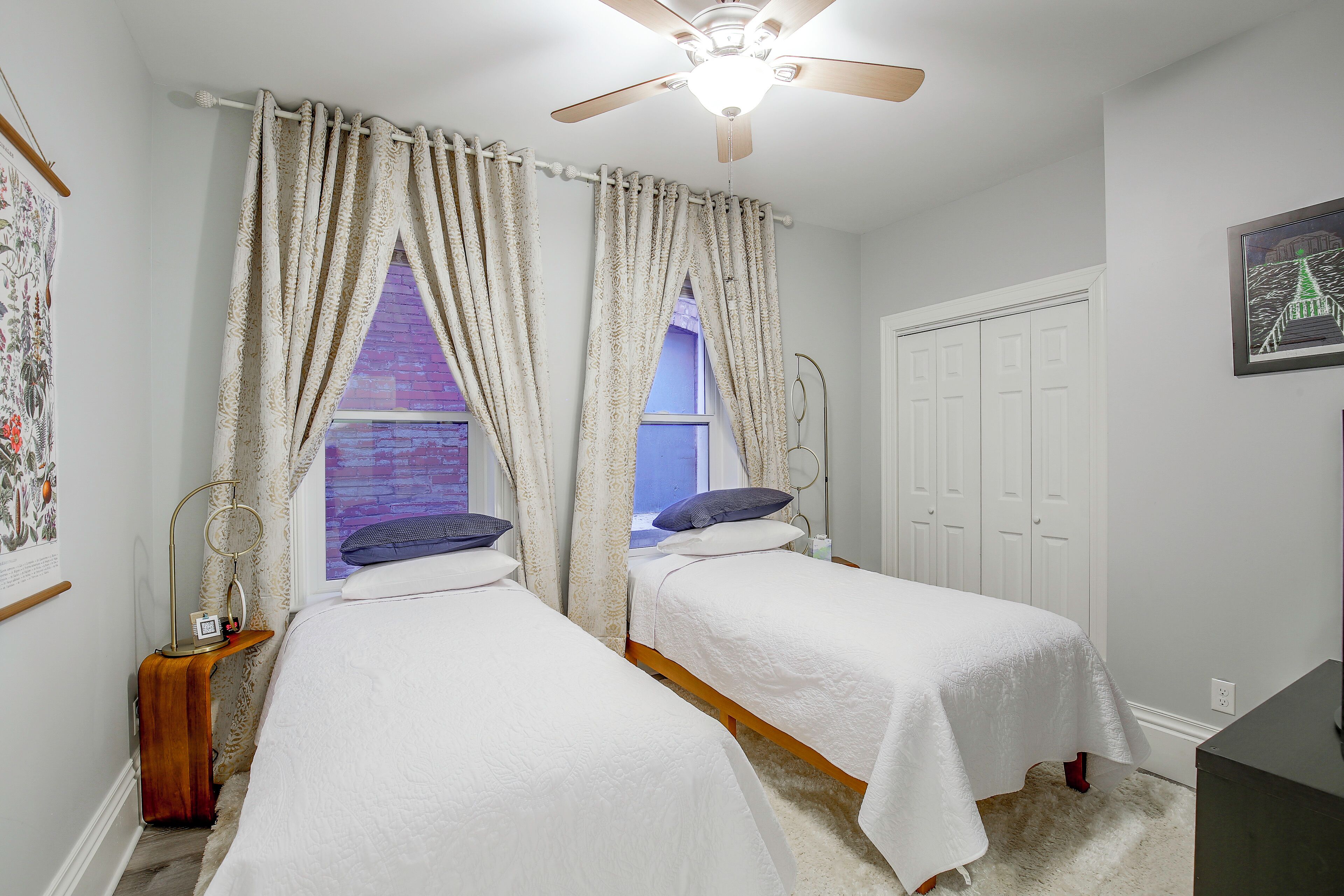 Quarto com duas camas de solteiro, um ventilador de teto, janela com cortinas, um quadro na parede e uma porta com espelho.