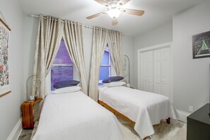 Quarto com duas camas de solteiro, um ventilador de teto, janela com cortinas, um quadro na parede e uma porta com espelho.