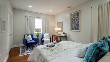Quarto de hotel com cama, área de estar com cadeiras, mesa com luminária, janela com cortinas e um quadro na parede.