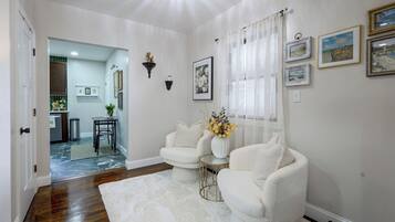 Quarto com tapete branco, duas poltronas brancas, uma mesa pequena e um vaso branco com flores. Há quadros nas paredes e uma porta que dá para outro cômodo.