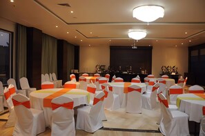 Uma sala de conferências com mesas e cadeiras redondas, um palco com um púlpito, e um lustre grande.