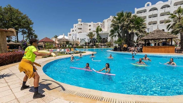 Área de piscina com várias pessoas, algumas em pranchas de surf, e uma pessoa vestida com um traje amarelo vibrante.