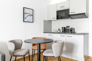 Uma cozinha moderna com armários brancos, um micro-ondas preto e uma máquina de café. Há um mapa emoldurado na parede e duas cadeiras ao redor de uma mesa pequena.