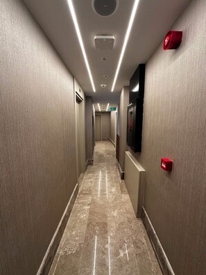 Corredor de hotel com piso de mármore, iluminação embutida, televisão de tela plana na parede e caixa de alarme de incêndio vermelha.
