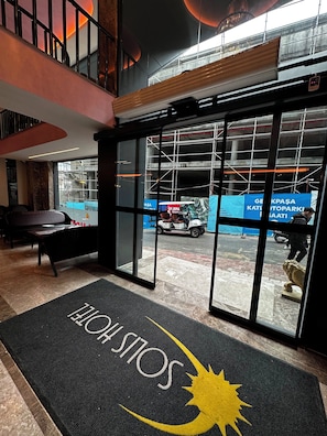 Entrada de hotel com tapete, balcão de recepção e vista da rua com obras.