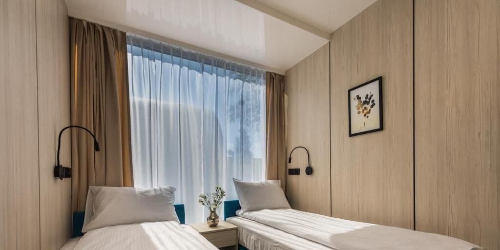 Quarto de hotel com duas camas, uma janela com cortinas leves, mesa de cabeceira com abajur e quadro na parede.