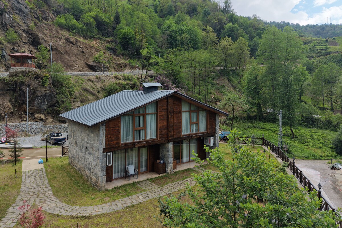 Uma casa de madeira de dois andares com base de pedra, um caminho de acesso pavimentado e vista para uma encosta arborizada.