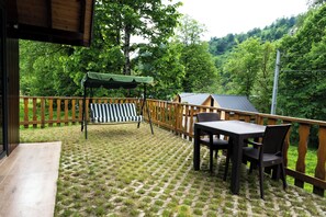 Um deck de madeira com mesa e cadeiras, uma cadeira balanço e vista para uma casa e floresta.