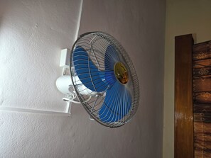 Um ventilador de parede com design azul e branco, posicionado em frente a uma parede lisa com um painel de madeira à direita.