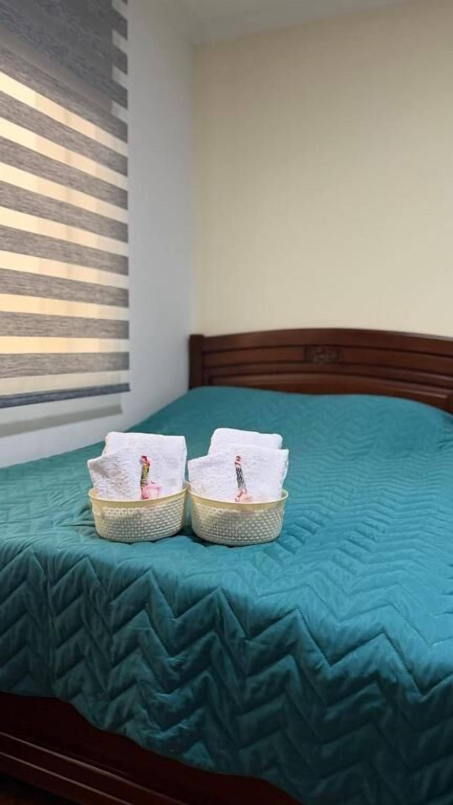 Uma cama com colcha verde-água, duas toalhas brancas em cestos e uma janela com persianas horizontais.