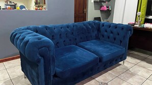 Um sofá azul com capitas num espaço com piso de azulejos, uma porta de madeira e uma cozinha com armários e uma janela.