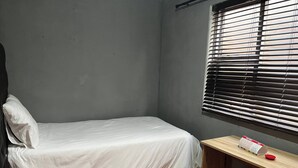 Quarto pequeno com uma cama de solteiro, piso de madeira, escrivaninha com cadeira e janela com persianas.