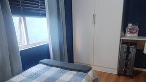 Quarto com cama, guarda-roupa, prateleira com artigos de higiene e janela com persianas.
