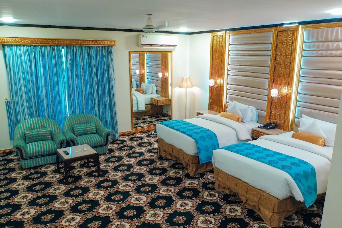Quarto de hotel com duas camas, área de estar com cadeiras, um espelho grande e um tapete estampado.