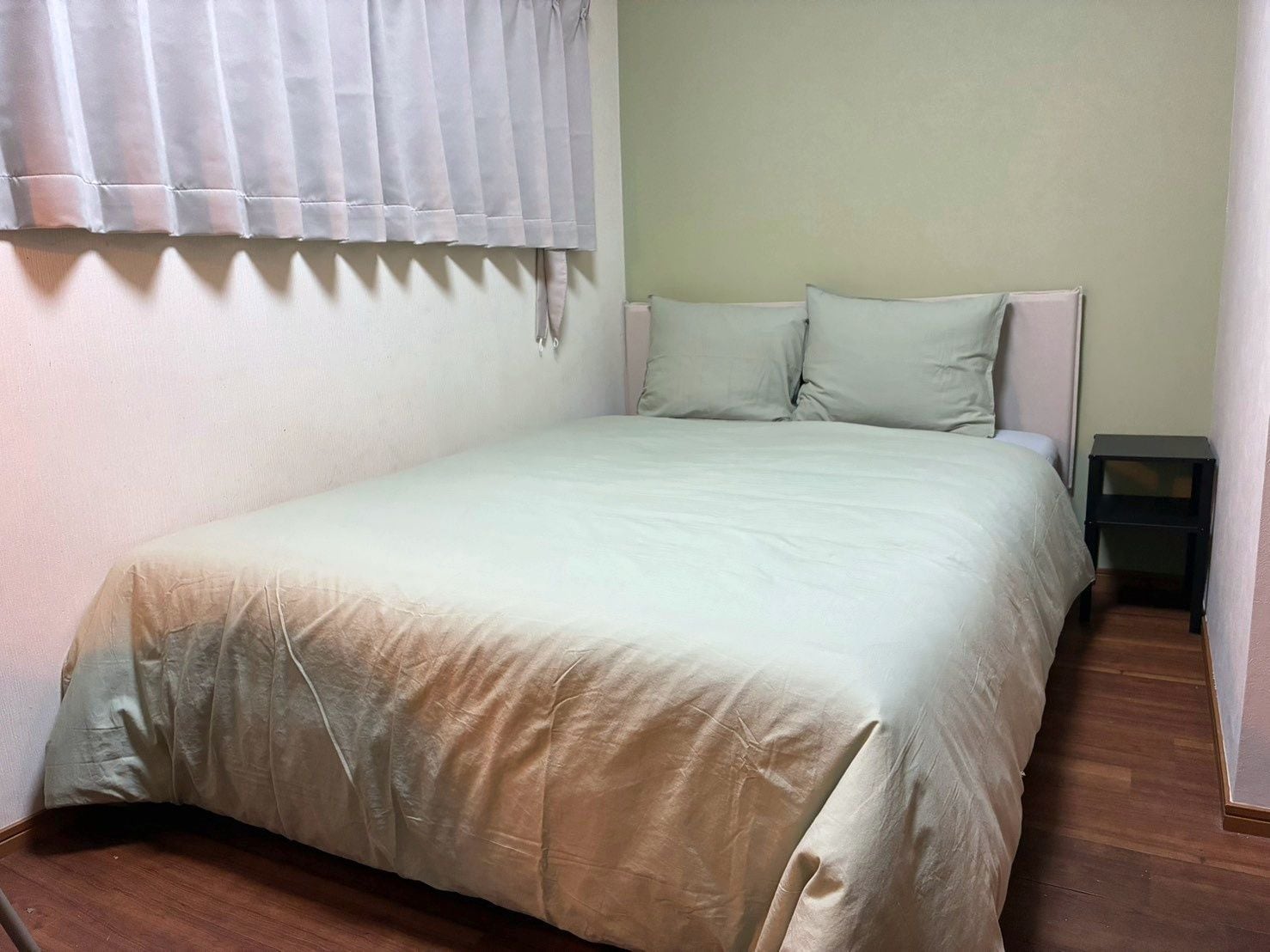 Uma cama de solteiro com um edredom claro em um quarto com piso de madeira e parede lisa.