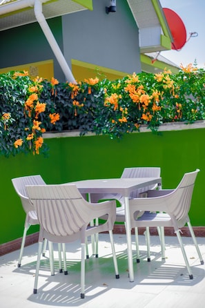 Uma área de estar externa com mesa e cadeiras, uma parede verde com flores laranja e uma antena parabólica vermelha.