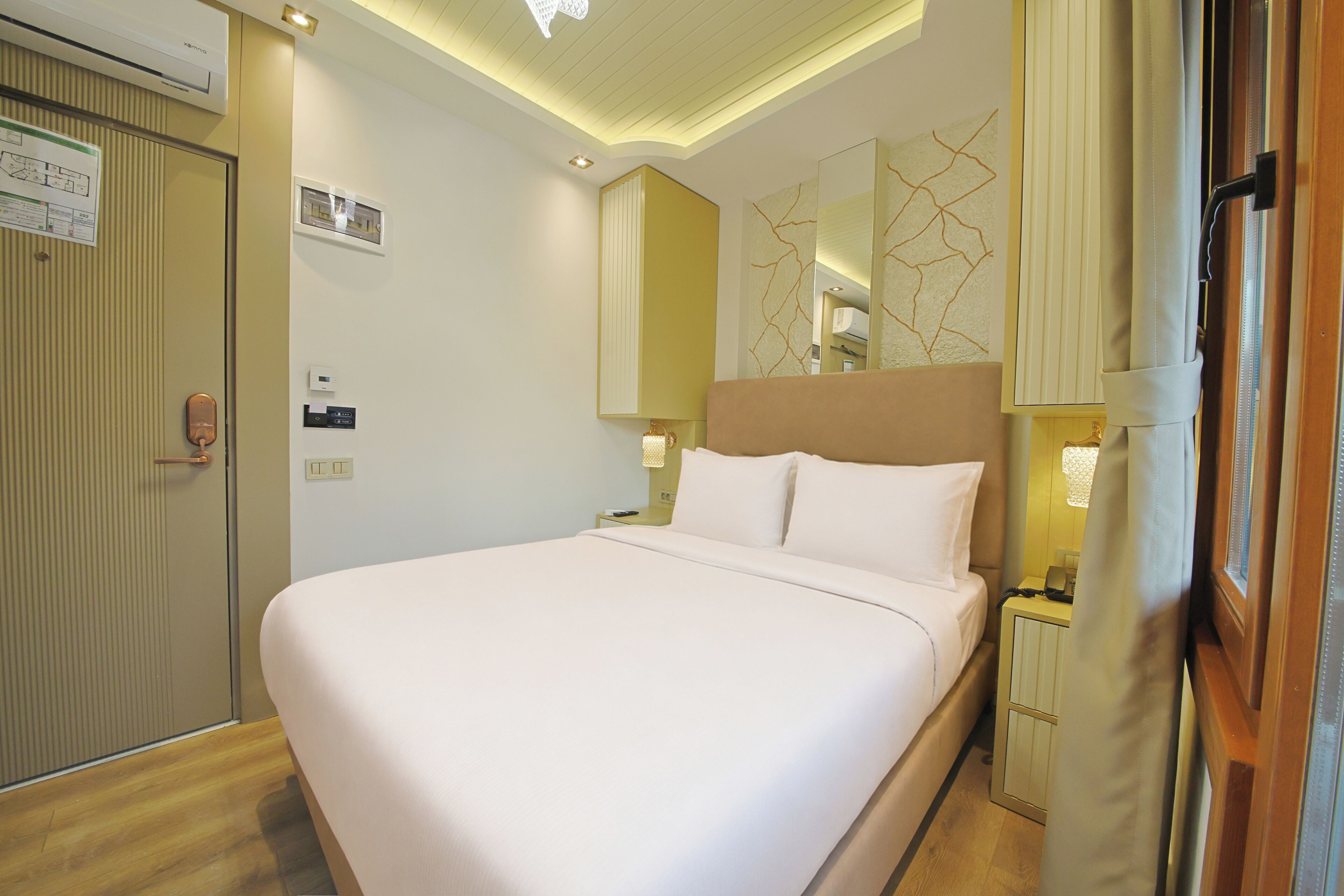 Quarto de hotel com uma cama grande, mesa de cabeceira, luminária, espelho e porta com fechadura.