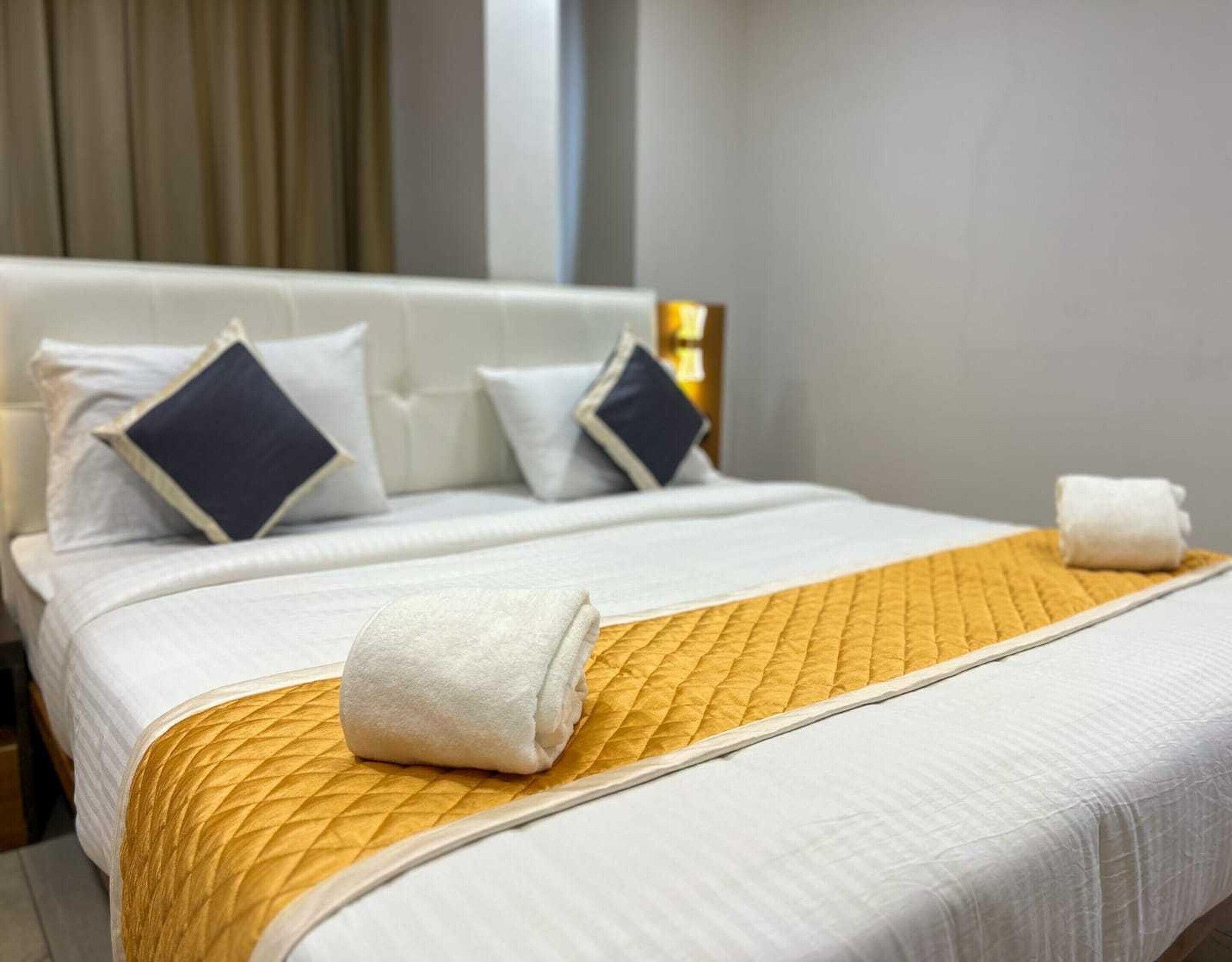 Quarto de hotel com cama arrumada, colcha dourada e duas almofadas com detalhes azul-escuros.