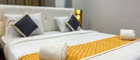 Quarto de hotel com cama arrumada, colcha dourada e duas almofadas com detalhes azul-escuros.