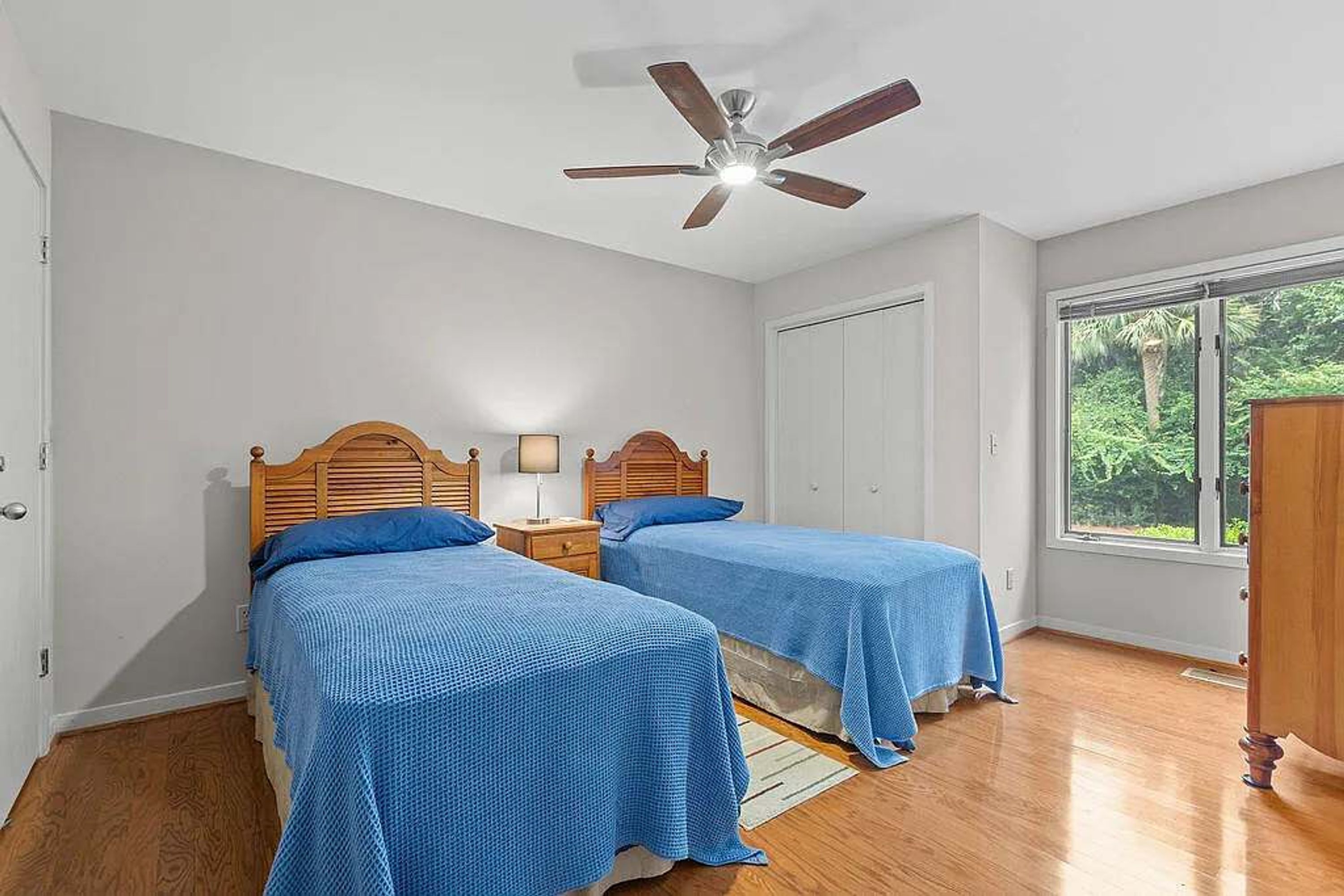 Quarto com duas camas, ventilador de teto, janela com vista para vegetação e uma porta.