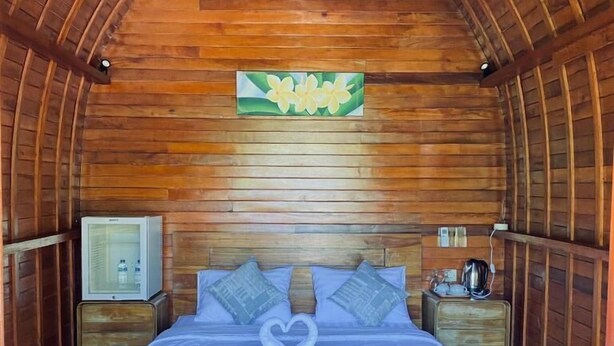Quarto de madeira com uma cama, uma mesinha de cabeceira, uma televisão e um candeeiro fixado na parede.