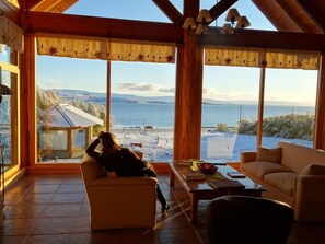 Quarto com grande janela com vista para uma praia e montanhas, mobiliado com sofá e mesa de centro.