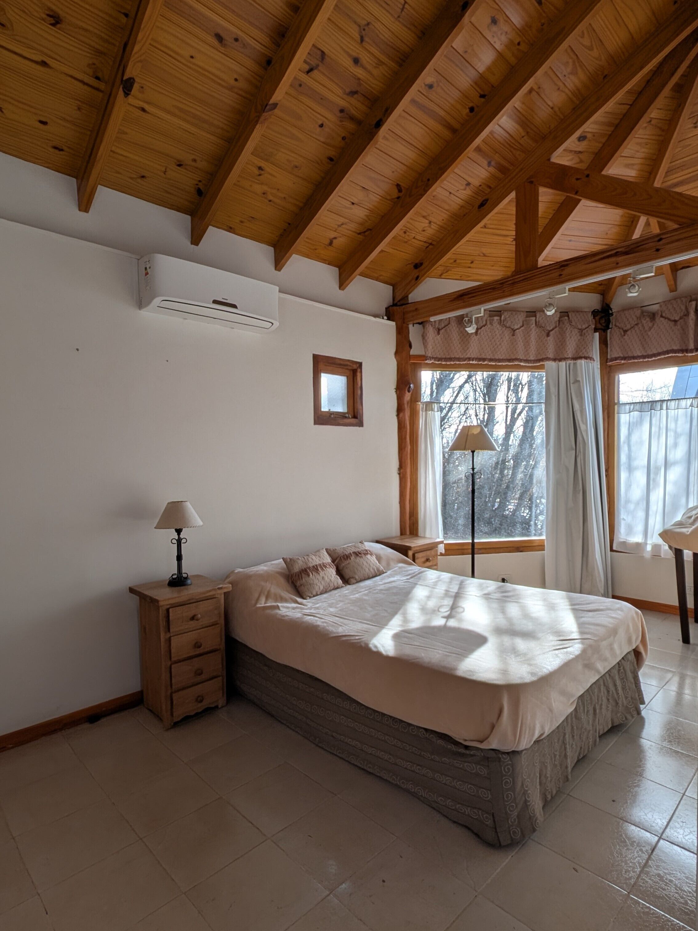 Quarto com teto de madeira, uma cama, mesa de cabeceira, abajur, janela com cortinas e um pequeno porta-retrato na parede.