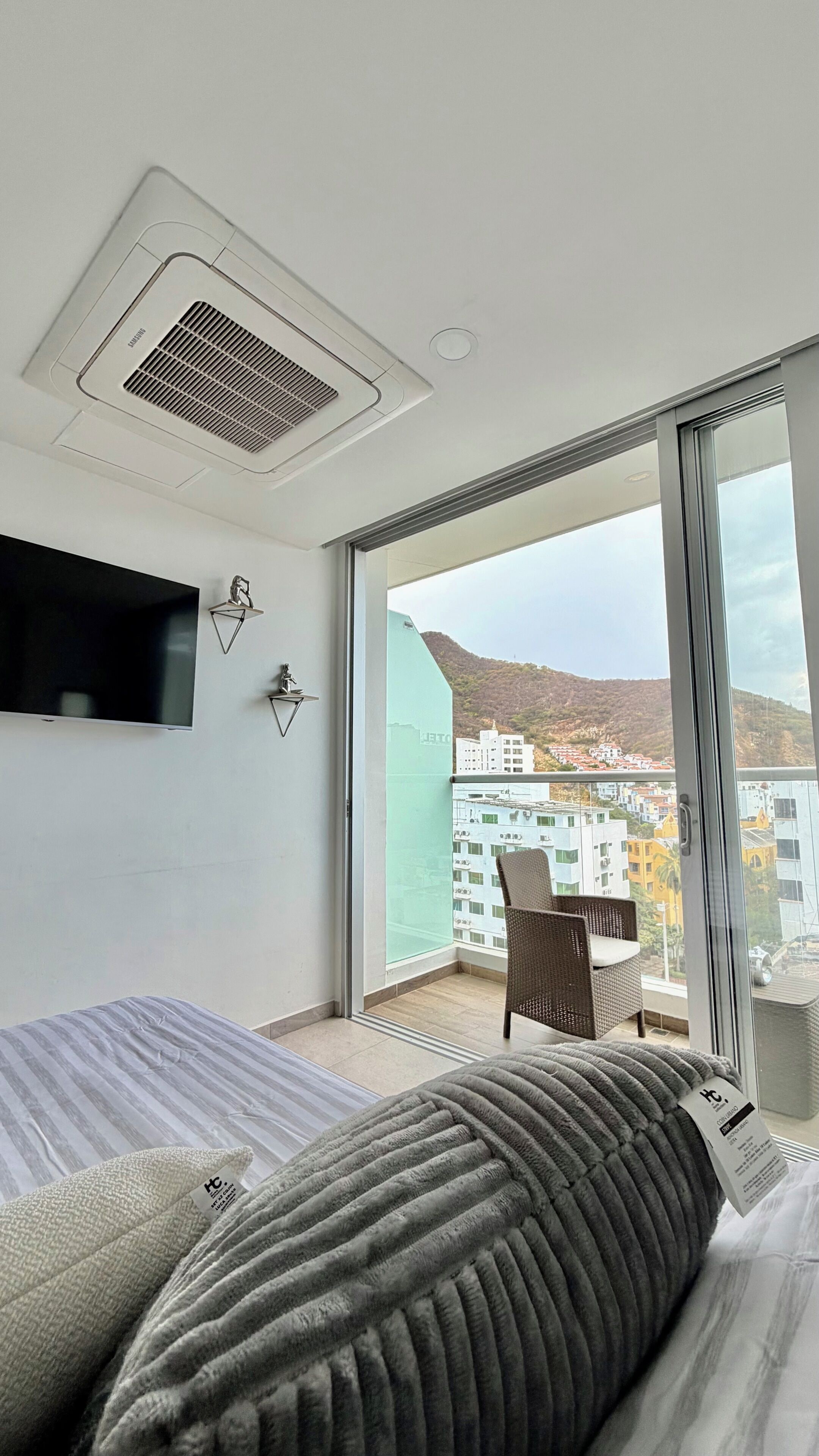 Quarto de hotel com cama, televisão, janela com vista para edifícios e montanha, e varanda com cadeira.