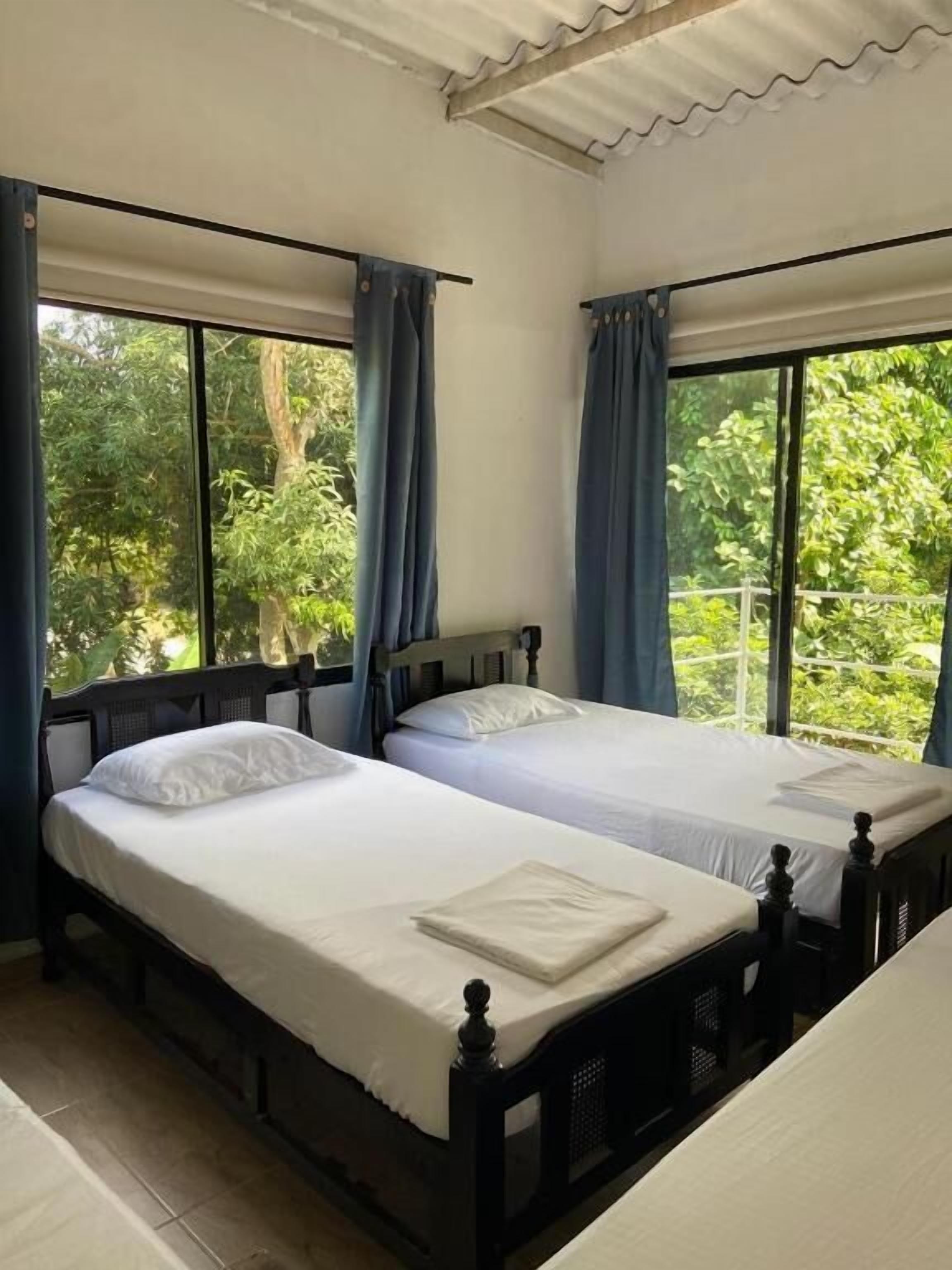Quarto com duas camas de solteiro, cada uma com cabeceira de madeira, roupa de cama branca e uma janela com cortinas azuis.