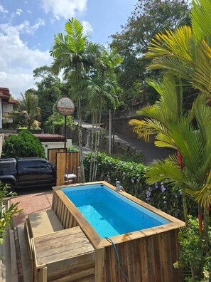 Piscina de madeira com superfície de água azul cristalina, cercada por vegetação exuberante e uma placa indicando uma área de quintal.
