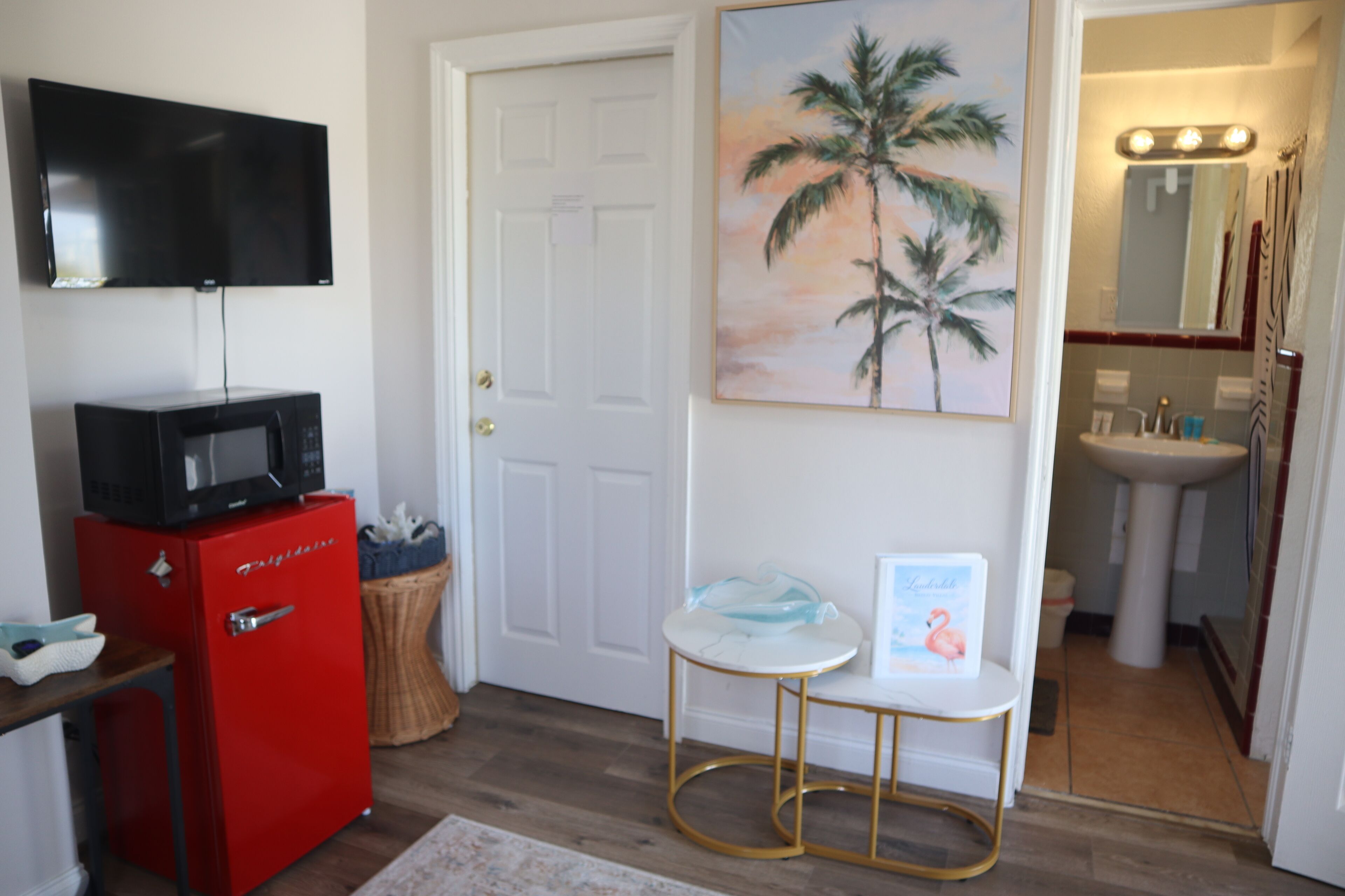 Quarto com televisão de tela plana na parede, um aparelho vermelho, uma moldura com imagem e uma mesa pequena com um objeto de vidro.