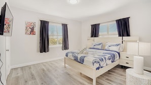 Quarto com cama, mesa de cabeceira, abajur e janela com cortinas.