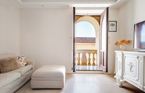 Quarto com sofá branco, um apoio para os pés combinando, armário branco e televisão. Uma porta aberta dá acesso a uma varanda externa com guarda-corrimão.