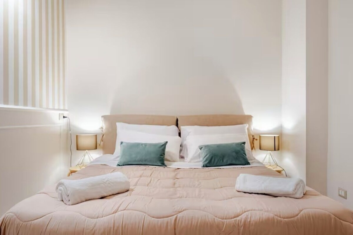 Cama bem arrumada com edredom bege e travesseiros, duas luminárias de mesa e uma janela com persianas verticais.