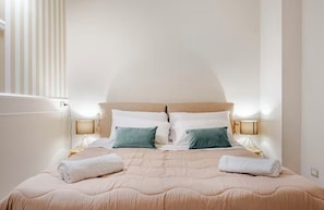 Cama bem arrumada com edredom bege e travesseiros, duas luminárias de mesa e uma janela com persianas verticais.
