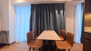 Espaço de refeições com mesa e cadeiras, janela com cortinas e pequeno espaço de cozinha.