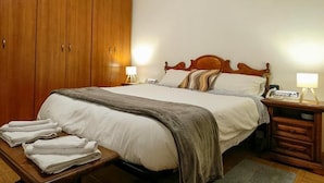 Quarto de hotel com um guarda-roupa de madeira grande, uma cama de solteiro com cabeceira de madeira, duas mesas de cabeceira com luminárias, e uma pequena mesa de cabeceira com um rádio.
