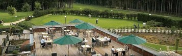 Uma área de refeições ao ar livre com mesas e cadeiras, rodeada por um deck de madeira e guarda-sóis verdes.