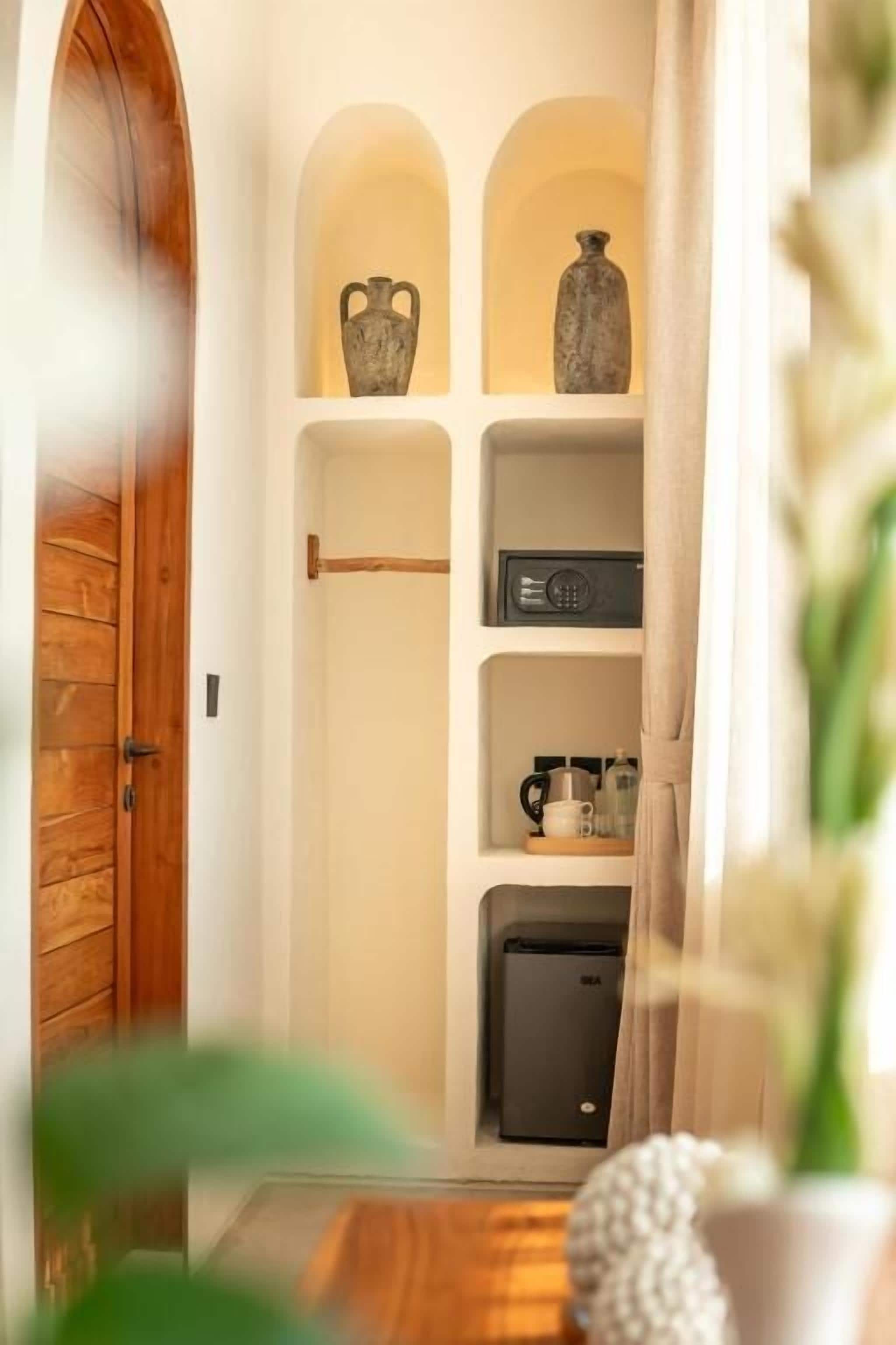 Um corredor com porta de madeira, prateleira com vasos decorativos, máquina de café e um pequeno frigorífico.