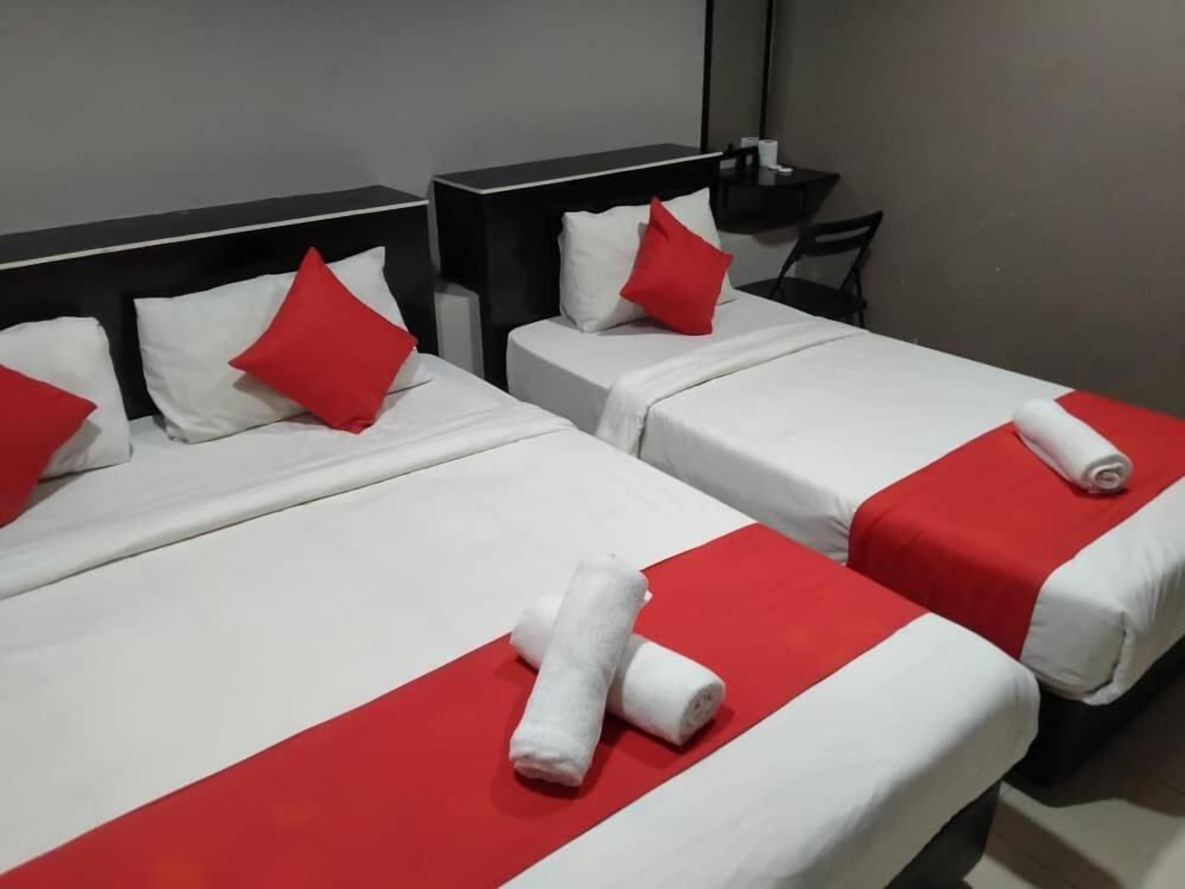 Quarto de hotel com duas camas, cada uma com roupa de cama branca e detalhes vermelhos, e uma pequena mesa de trabalho com uma cadeira.