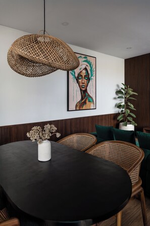 Uma sala de jantar com uma mesa redonda, cadeiras de vime, luminária pendente, obra de arte emoldurada e um vaso com planta.