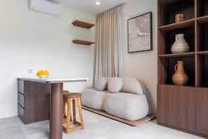 Um espaço de estar moderno com um sofá, uma mesinha, um banquinho, uma estante com objetos decorativos e um ar condicionado fixado na parede.