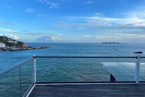 Vista do convés de um navio, com grades, mar calmo e terra distante com edifícios.